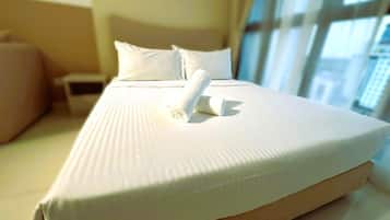 Uma cama bem arrumada com roupa de cama branca e uma toalha enrolada, uma mesa de cabeceira e uma janela com vista para edifícios.