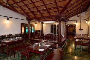 Uma área de refeições com mesas e bancos de madeira, teto alto com vigas aparentes e lustres pendurados.