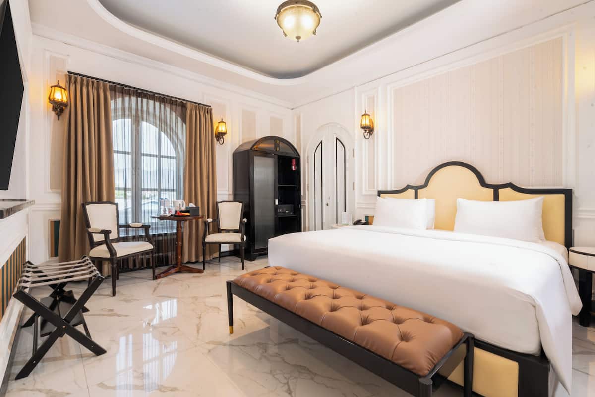 Quarto de hotel com uma cama grande, uma escrivaninha com cadeira, um banco e um guarda-roupa.