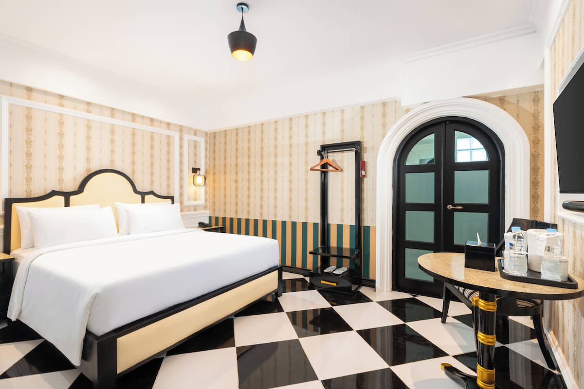 Quarto de hotel com cama, mesa redonda com amenidades, cabideiro e porta com painel de vidro.