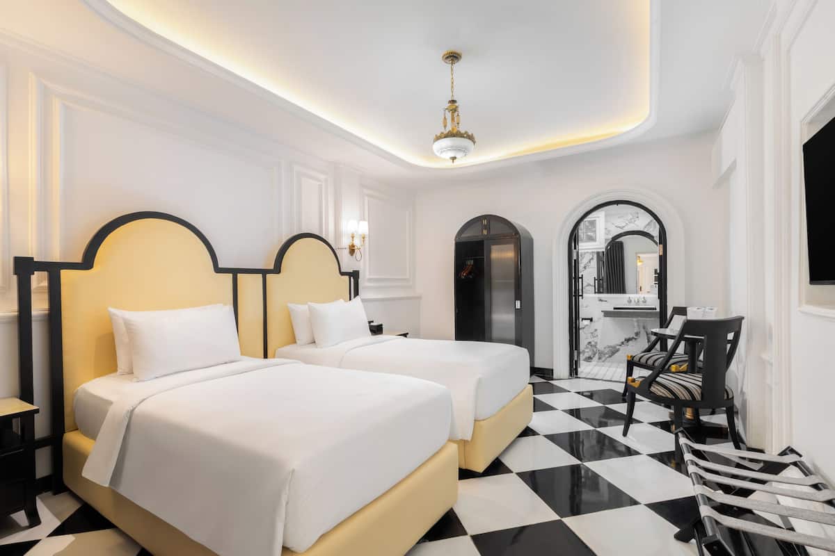 Quarto de hotel com duas camas, piso xadrez, lustre e espelho.