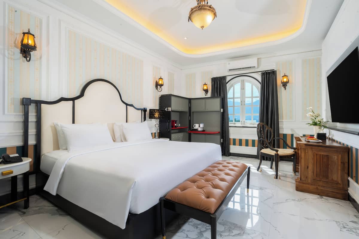 Quarto de hotel com uma cama grande, um banco de madeira, uma mesa e vista para o exterior.