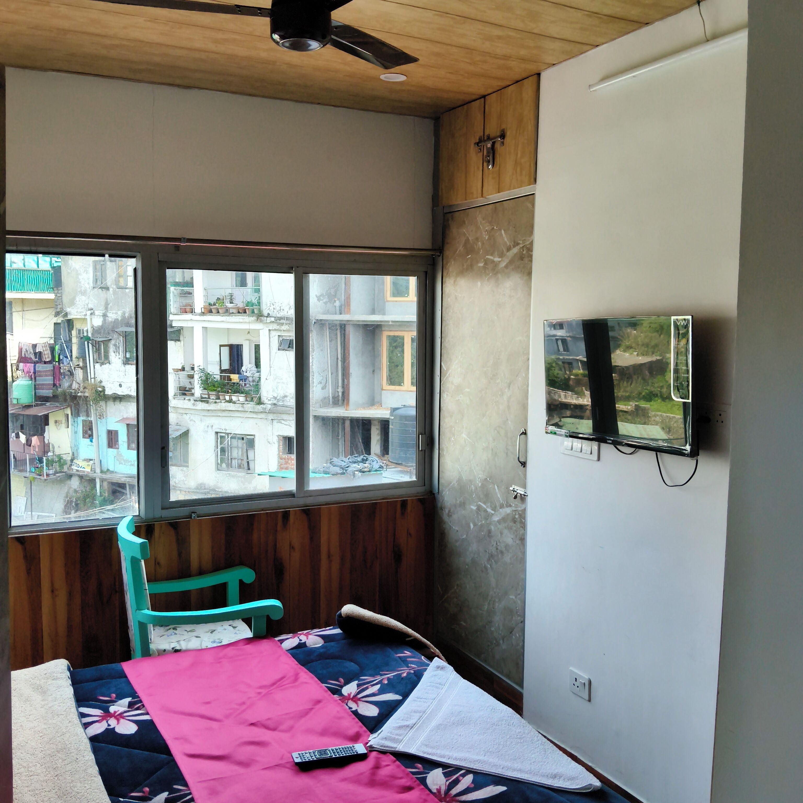Quarto com cama, televisão na parede e janela com vista para uma área residencial.