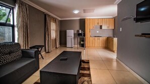 Uma sala de estar moderna com um sofá, uma mesa de centro e uma televisão. Há uma área de cozinha com armários de madeira e uma geladeira.
