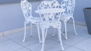 Uma área externa com assentos brancos, uma mesa, cadeiras, uma janela com cortinas e um vaso de planta.