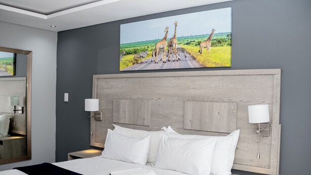 Quarto de hotel com cama grande, roupa de cama branca, cabeceira de madeira, duas luminárias de apoio e um quadro com girafas e zebras na parede.