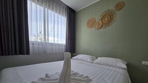 Quarto de hotel com uma cama grande, janela com cortinas leves e painel decorativo de artebricolagem na parede.