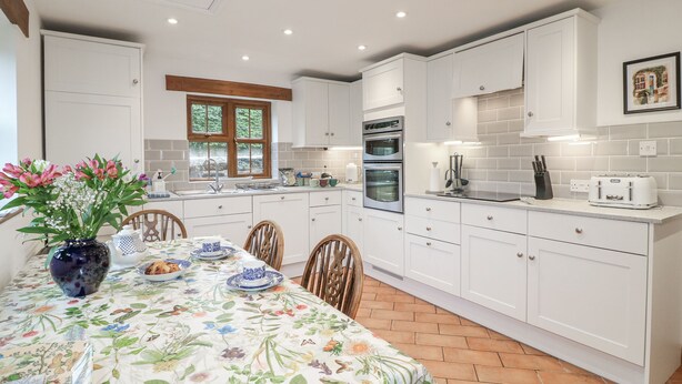 Cozinha moderna com armários brancos, forno duplo e área de jantar com toalha de mesa florida.
