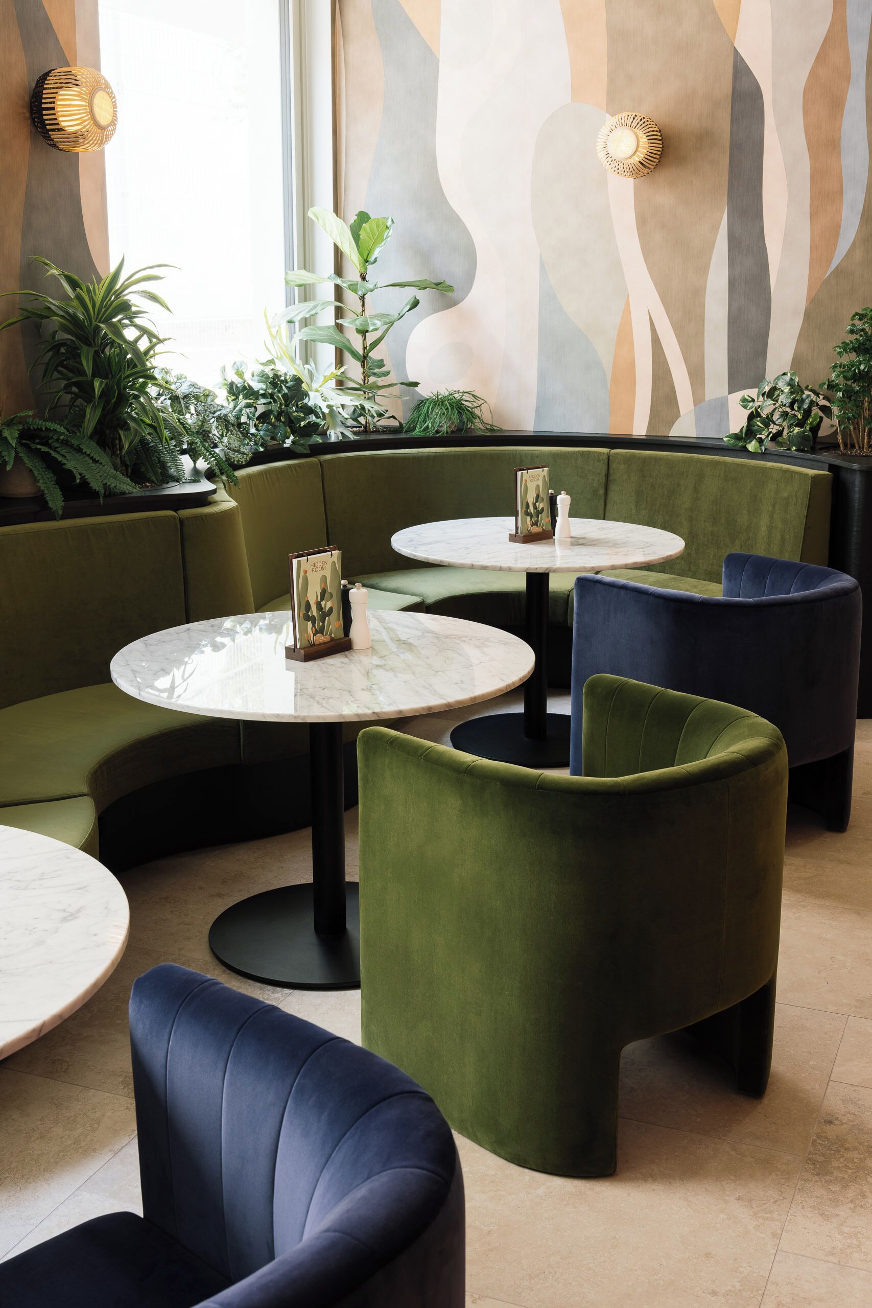 Uma área de estar com mesas redondas e cadeiras acolchoadas, parede com padrão decorativo e plantas em vasos.