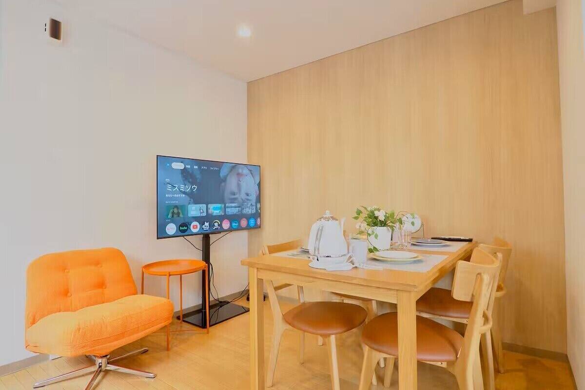Quarto moderno com mesa de jantar de madeira, TV fixada na parede e uma cadeira laranja vibrante.