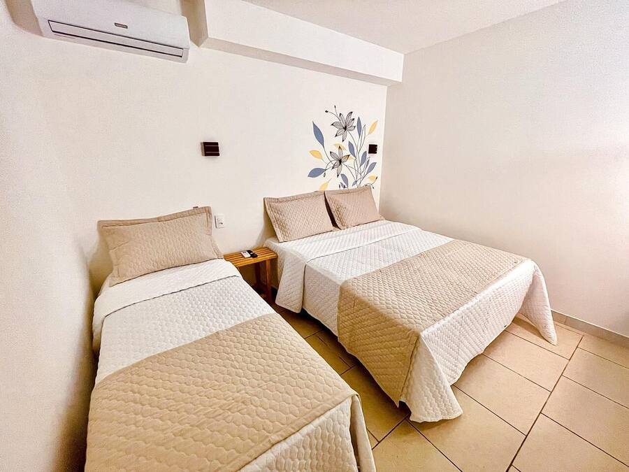 Quarto com duas camas, cada uma com colcha acolchoada bege e almofadas a combinar. Há uma pequena mesa de cabeceira de madeira entre as camas, e um ar condicionado fixado na parede acima das camas.
