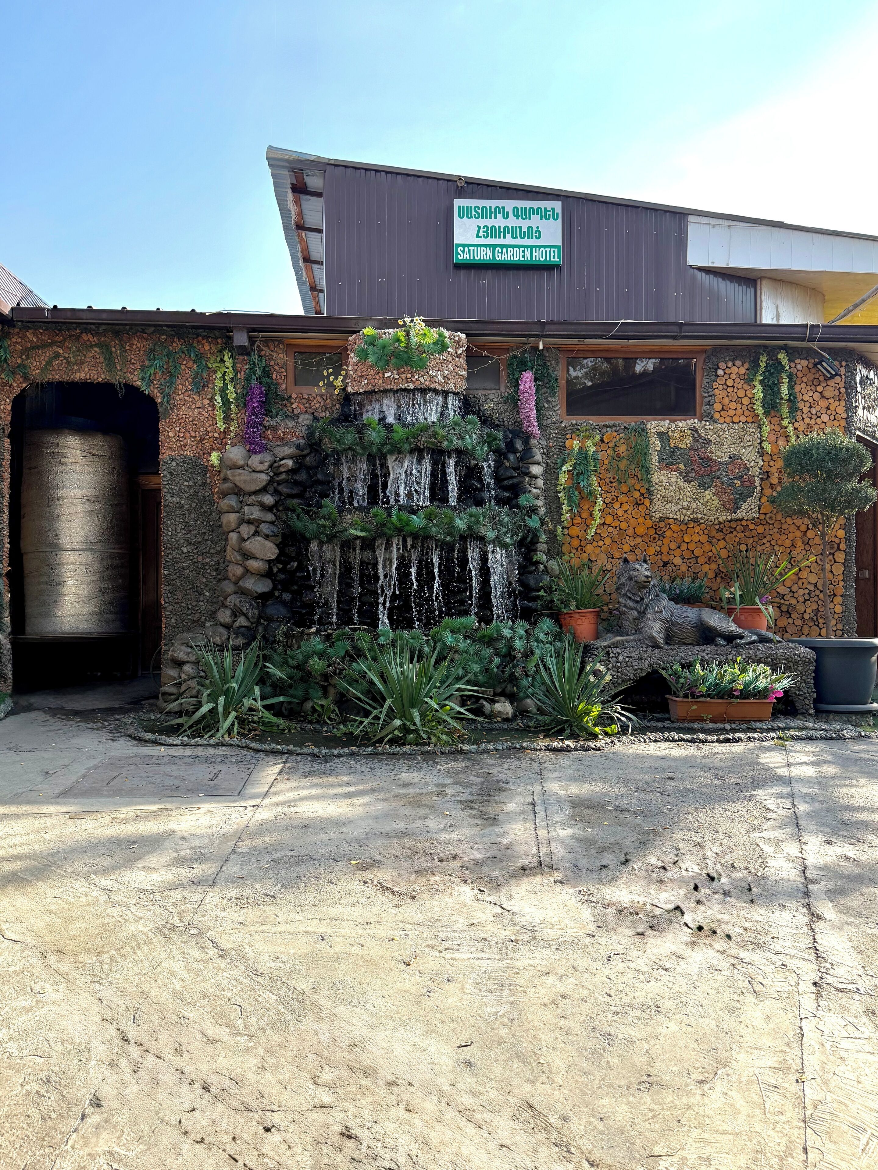 Entrada de hotel com parede de pedra, cascata e plantas em vasos.
