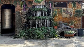 Entrada de hotel com parede de pedra, cascata e plantas em vasos.