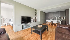 Uma sala de estar moderna com televisão de tela plana, uma mesa de centro e um sofá. Há uma área de jantar com uma mesa e cadeiras. O cômodo possui piso de madeira e paredes em tons claros.