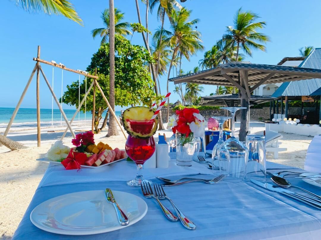 Disposição para refeições à beira-mar, com uma mesa posta para dois, apresentando uma bebida tropical, um prato de frutas e flores.