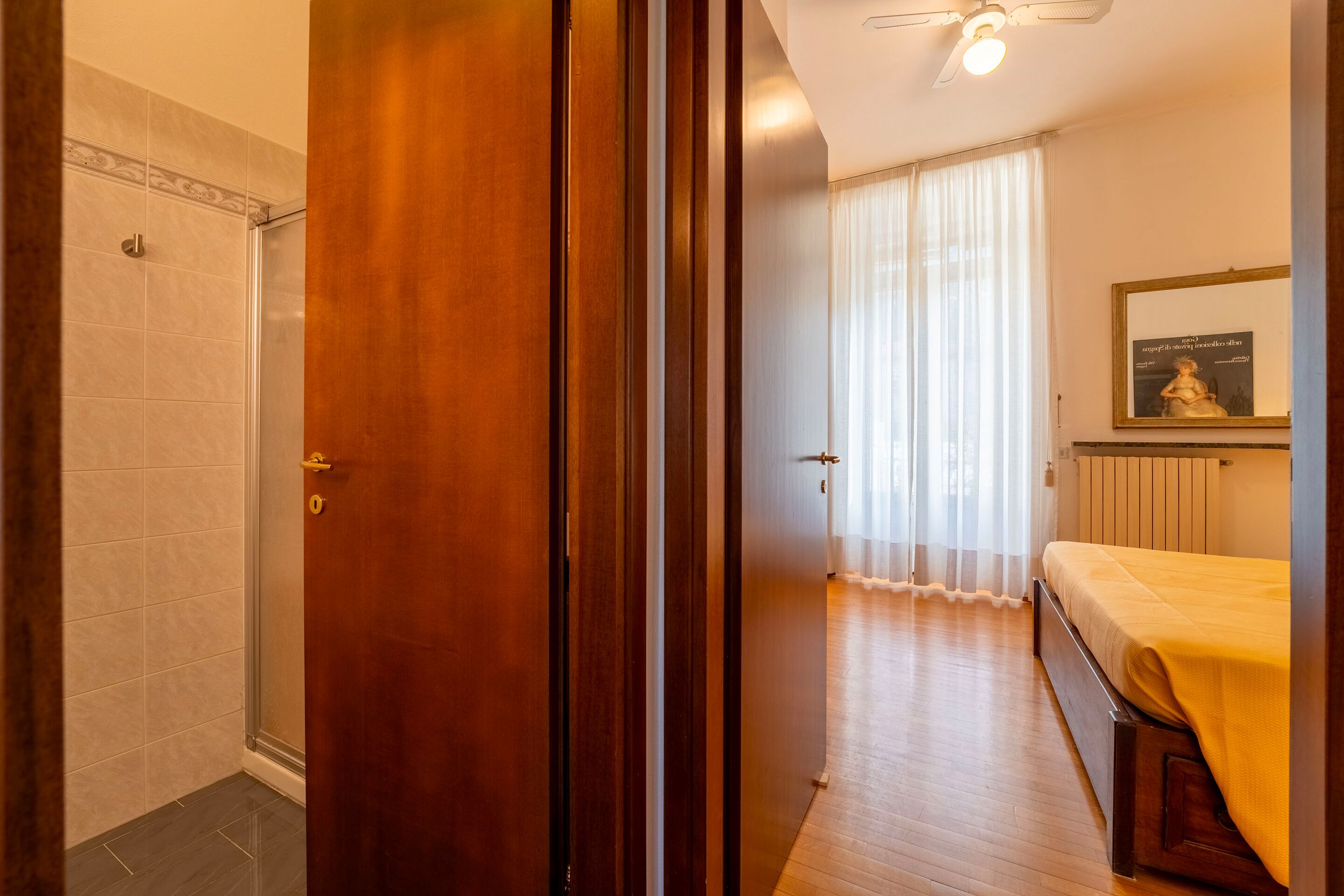 Quarto de hotel com porta de madeira, área de chuveiro, cama com colcha amarela e janela com cortinas translúcidas.
