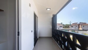 Varanda estreita com guarda-corrimão preto, uma porta à esquerda e vista ampla de edifícios ao fundo.