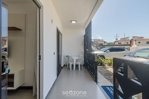 Uma varanda estreita com mesa e cadeiras, uma porta de correr e vista para carros estacionados e edifícios ao fundo.