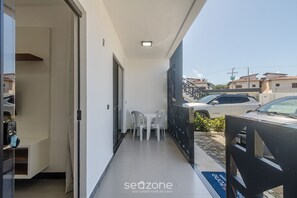 Uma varanda estreita com mesa e cadeiras, uma porta de correr e vista para carros estacionados e edifícios ao fundo.
