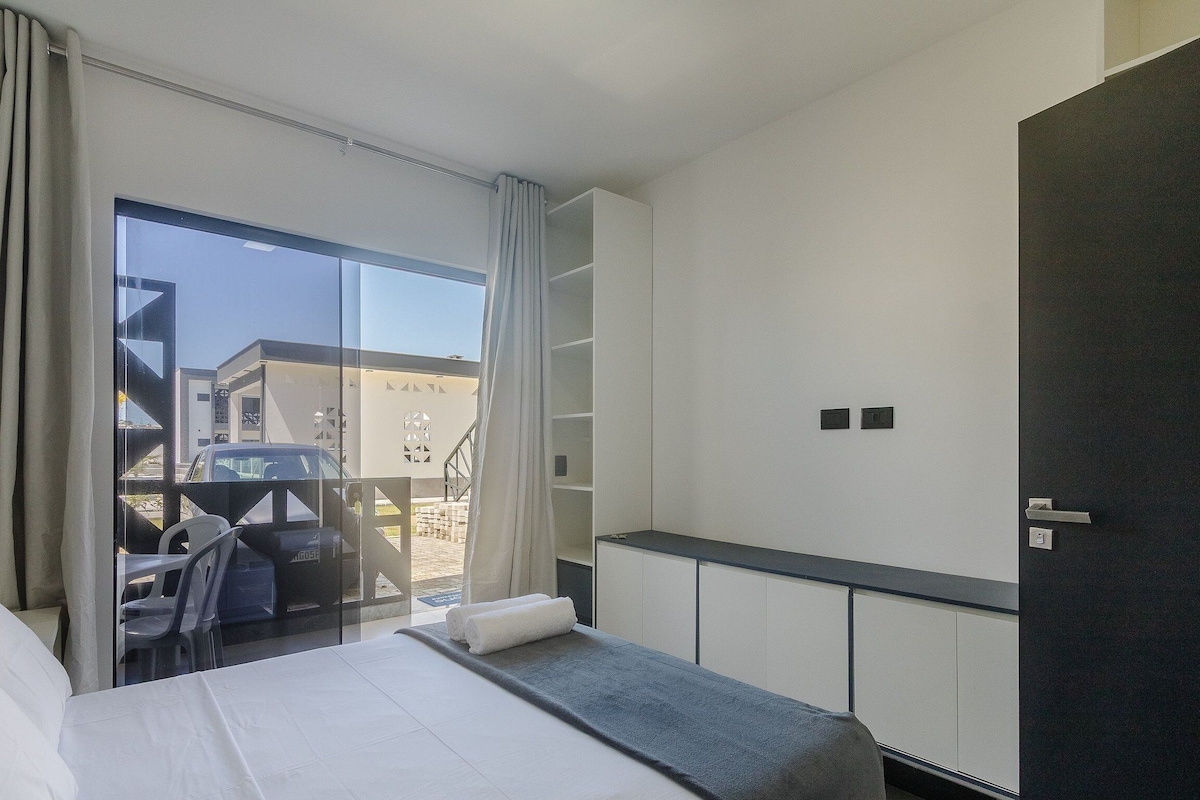 Quarto de hotel com cama, uma pequena mesa, área de estar com cadeiras, uma janela grande e vista para o exterior.