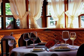 Mesa posta com copos de vinho, pratos de comida e uma vela, com cortinas e janelas ao fundo.