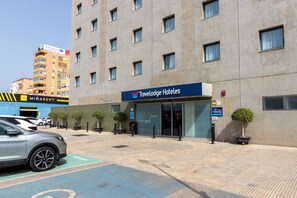 Um hotel Travelodge com fachada moderna, área de estacionamento e um edifício ao fundo.