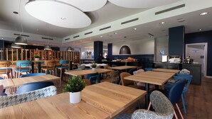 Restaurante moderno com mesas de madeira e cadeiras azuis, área de cozinha e um espaço de estar com um banco estampado.