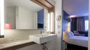Casa de banho de hotel com uma pia, espelho e dois secadores de cabelo. Há uma cama com um travesseiro e uma mesa de cabeceira com uma lâmpada.