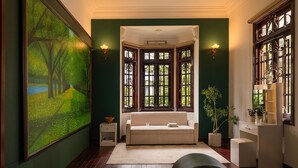Quarto com uma grande pintura de um parque, um parapeito com vários painéis, um sofá, uma mesa pequena e um armário com espelho.