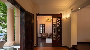 Um salão imponente com piso de madeira, uma grande porta de madeira escura e vista para um ambiente com lustre e sofá.