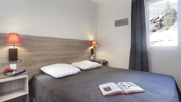 Cama bem arrumada com colcha xadrez vermelha e cinza, duas almofadas brancas e um livro aberto com óculos por cima. Há duas luminárias de cabeceira com abajures vermelhos, uma cabeceira de madeira e uma janela com vista para um prédio e árvores.