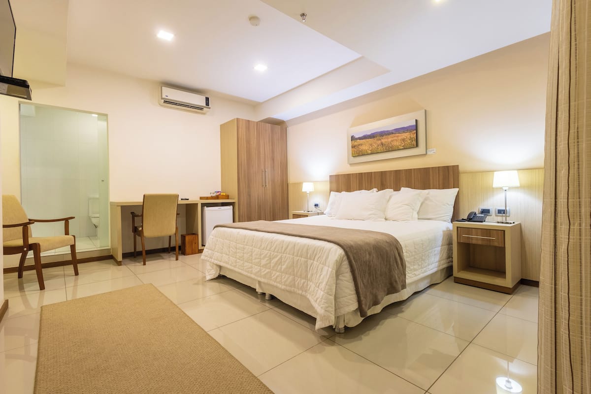 Quarto de hotel com uma cama grande, uma escrivaninha, uma cadeira, uma mesa de cabeceira e um quadro na parede.