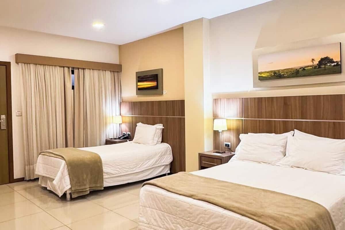 Quarto de hotel com duas camas, mesa de cabeceira, luminária, quadro na parede e janela com cortinas.