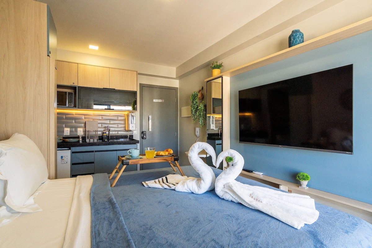Quarto de hotel moderno com cama, televisão, cozinha compacta e área de refeições.