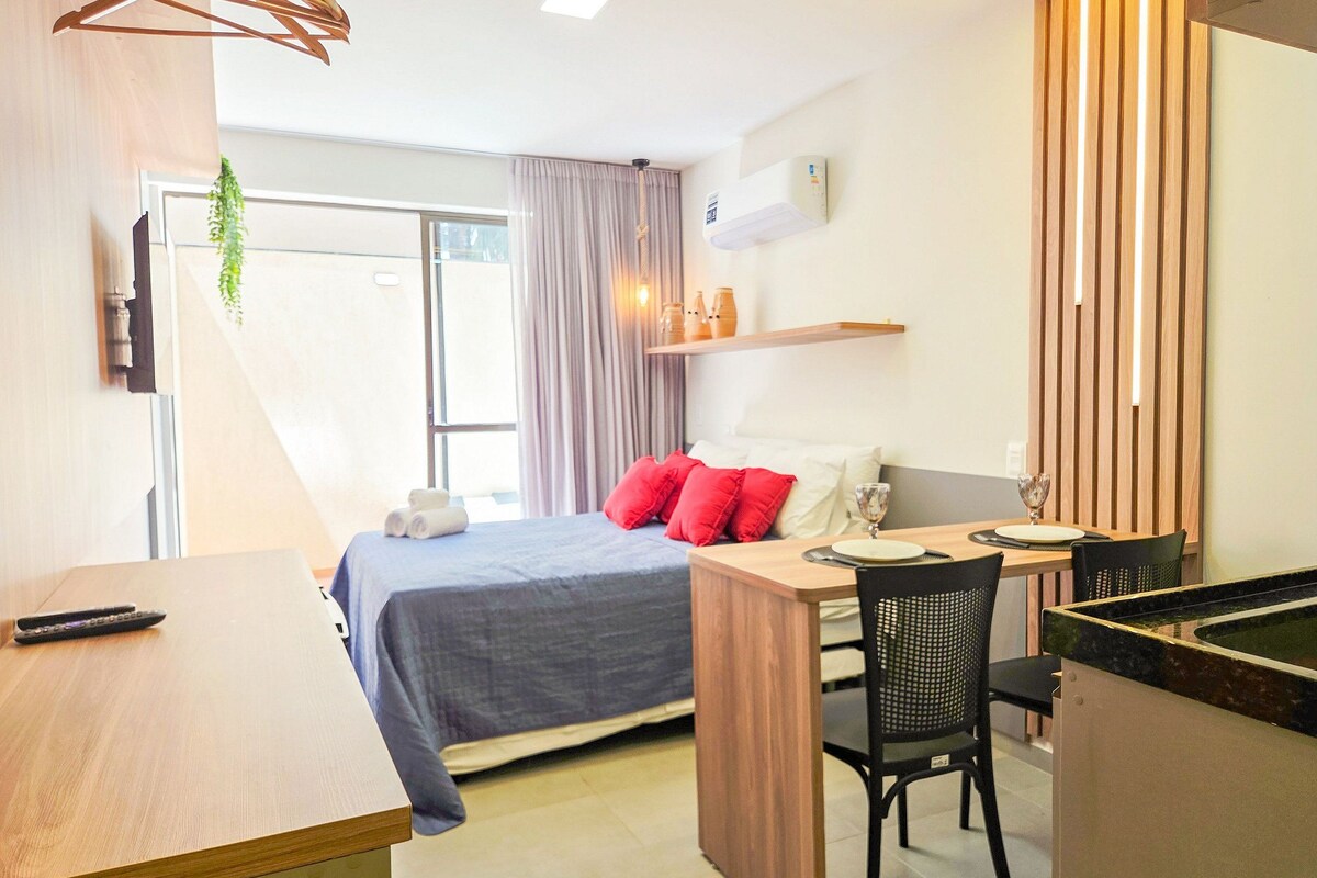 Quarto de hotel compacto com cama, escrivaninha, cadeira e pequeno espaço de cozinha.