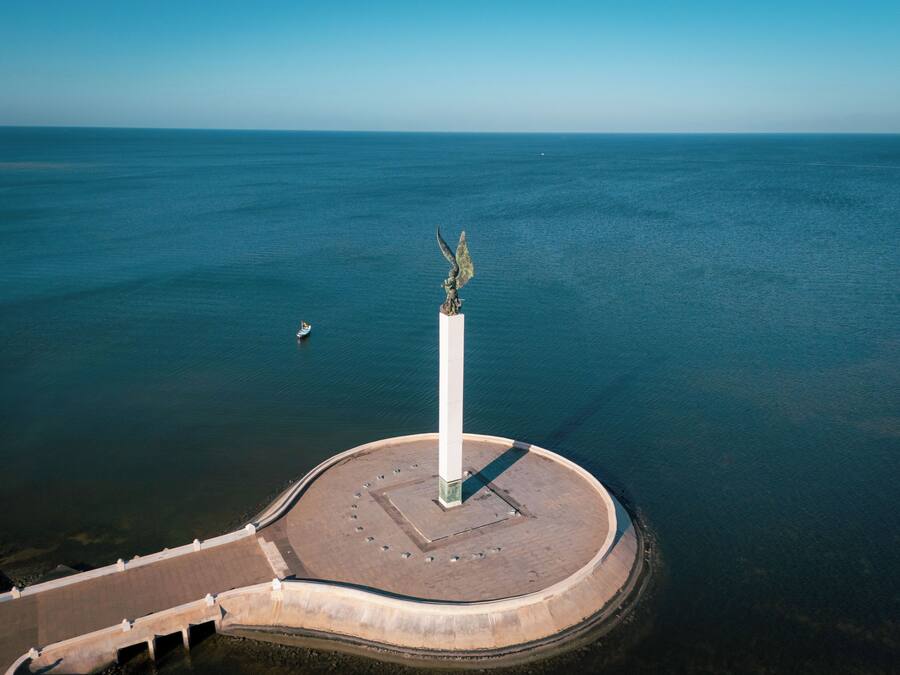 Vista aérea de uma orla marítima com um monumento central, uma praça circular e um mar calmo.