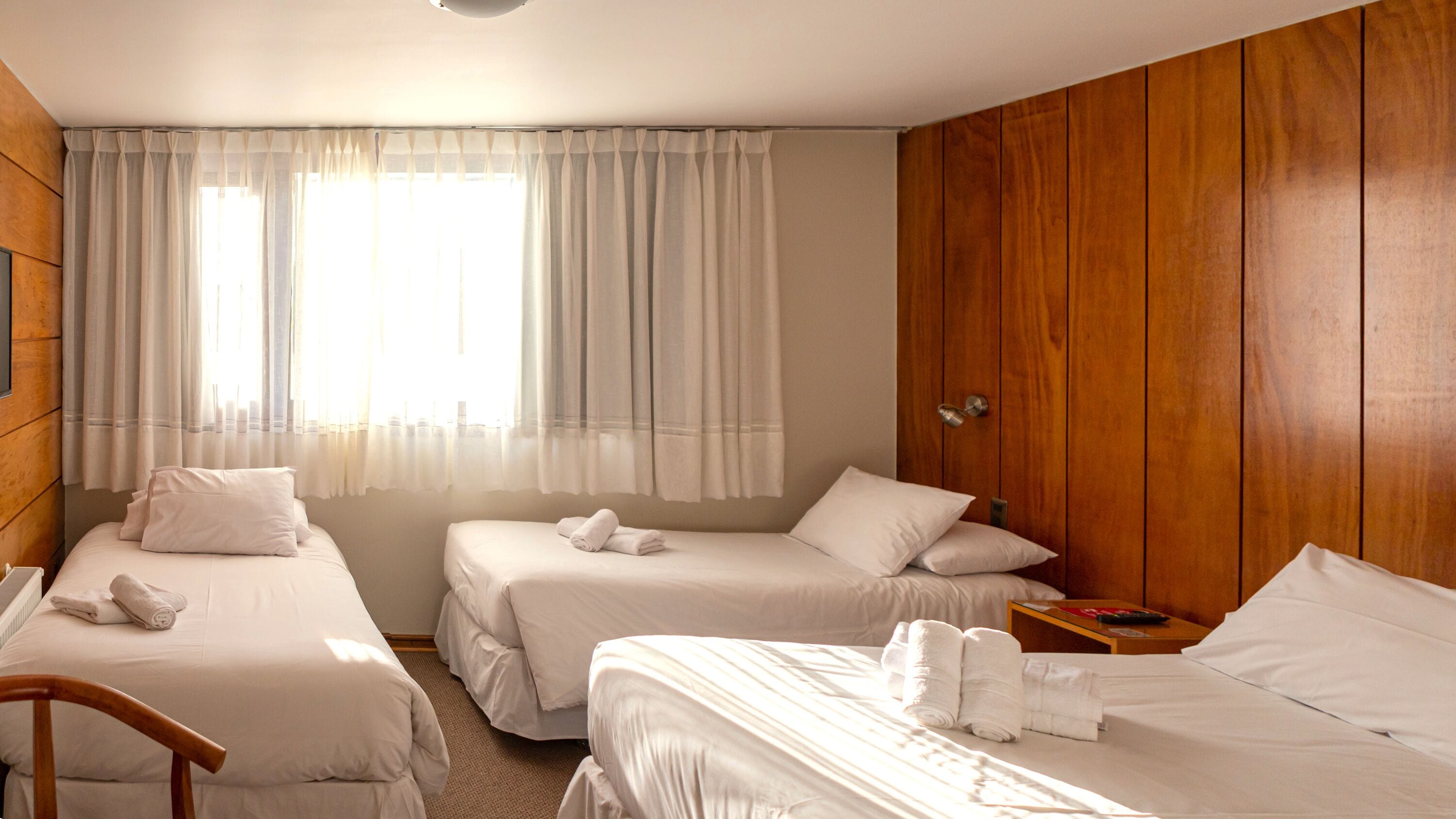 Quarto de hotel com duas camas de solteiro, um guarda-roupa de madeira, uma mesa pequena e uma janela com cortinas leves.