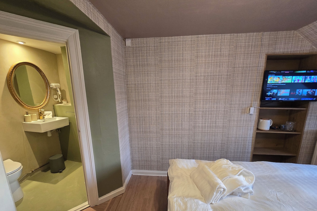Quarto de hotel com cama, televisão, banheiro com espelho e prateleira com objetos.