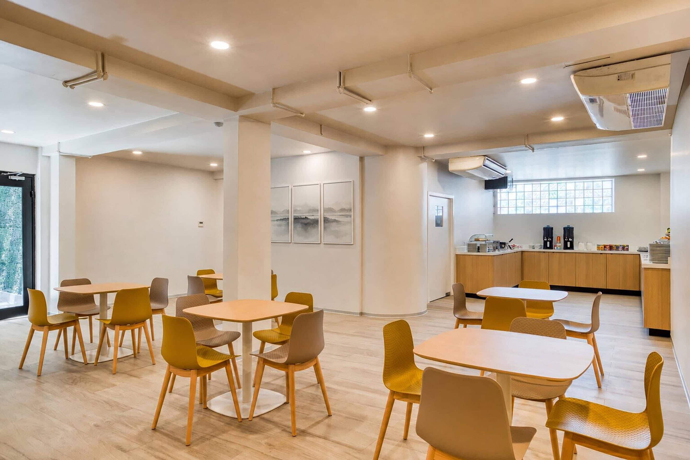 Uma sala de jantar moderna com mesas redondas e cadeiras amarelas, uma cozinha ao fundo e uma grande janela que ilumina naturalmente o espaço.