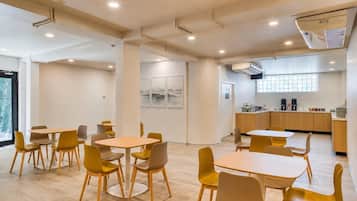 Uma sala de jantar moderna com mesas redondas e cadeiras amarelas, uma cozinha ao fundo e uma grande janela que ilumina naturalmente o espaço.