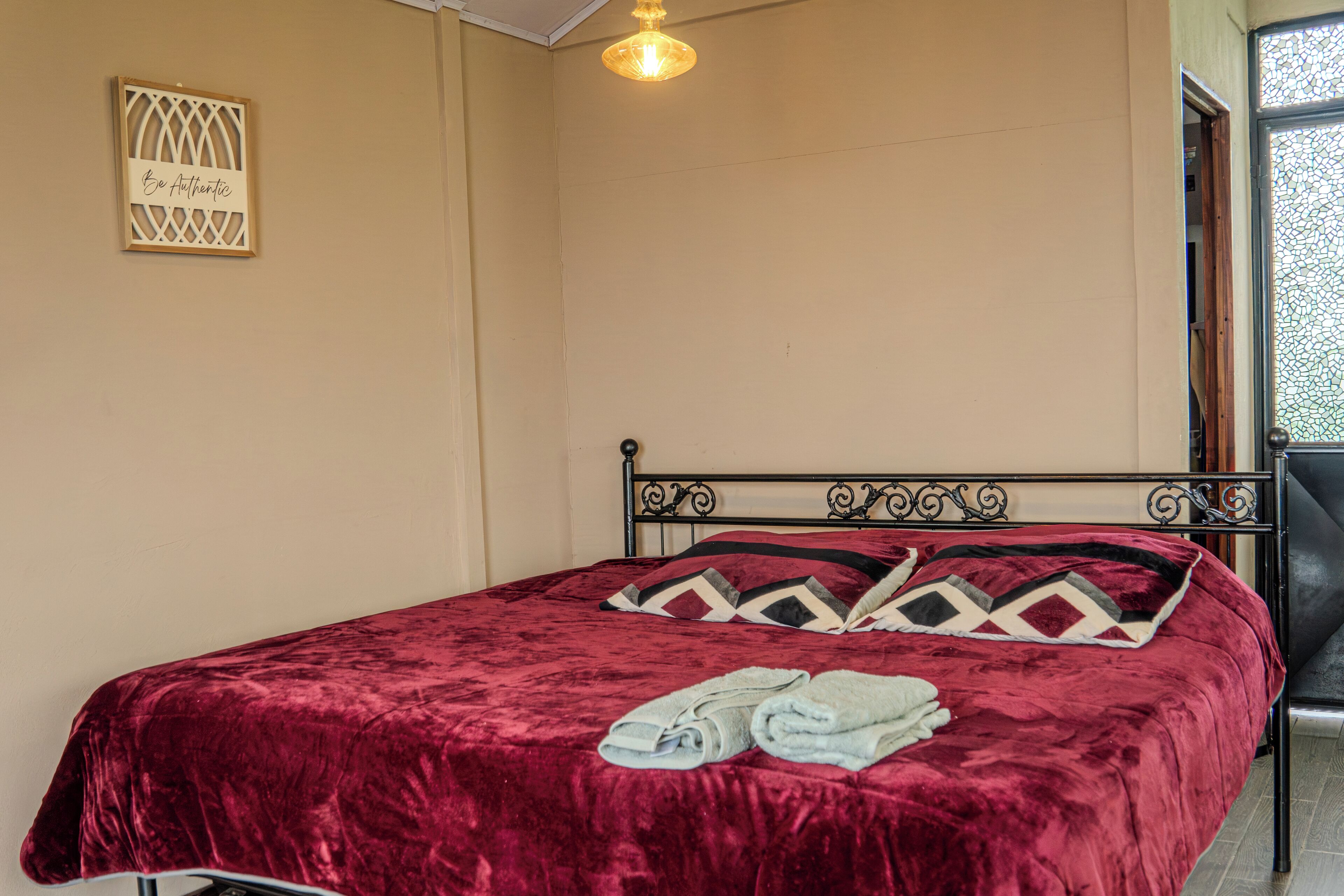 Uma cama bem arrumada com colcha e travesseiros vermelhos, um quadro na parede e uma janela com vista.