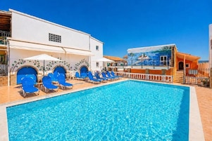 Uma piscina com cadeiras azuis, um edifício branco com arcos e um mural no edifício vizinho.
