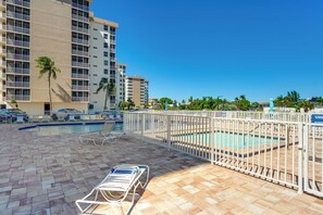 Uma área de piscina com espreguiçadeiras, piscina e um edifício com várias varandas ao fundo.
