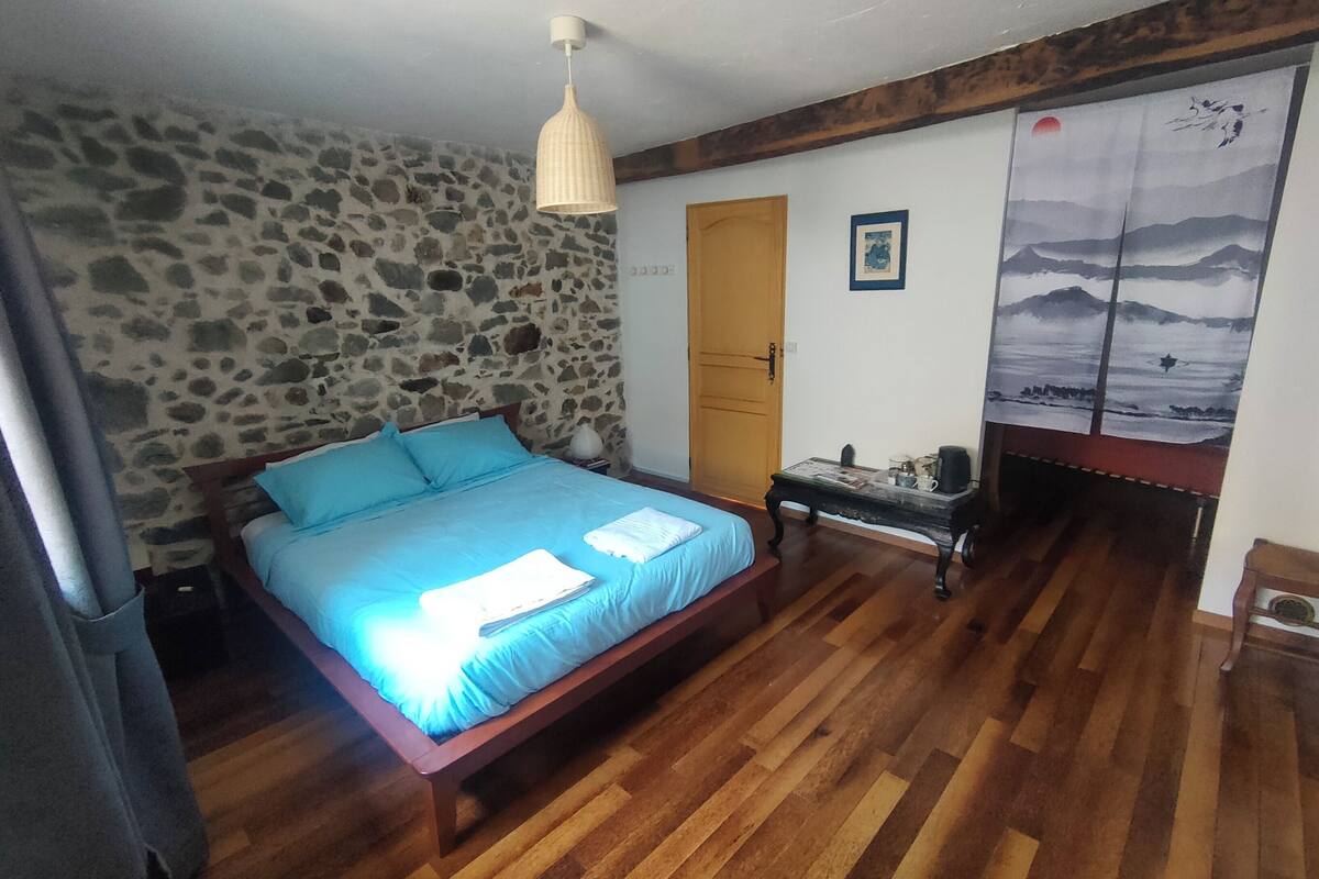 Quarto com parede de pedra, cama de madeira, piso de madeira, luminária pendente, porta de madeira e quadro emoldurado.