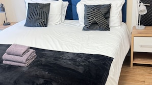 Cama bem arrumada com cabeceira azul, roupa de cama branca e almofadas estampadas. Uma toalha dobrada está sobre a cama, e uma cadeira verde é visível em primeiro plano.