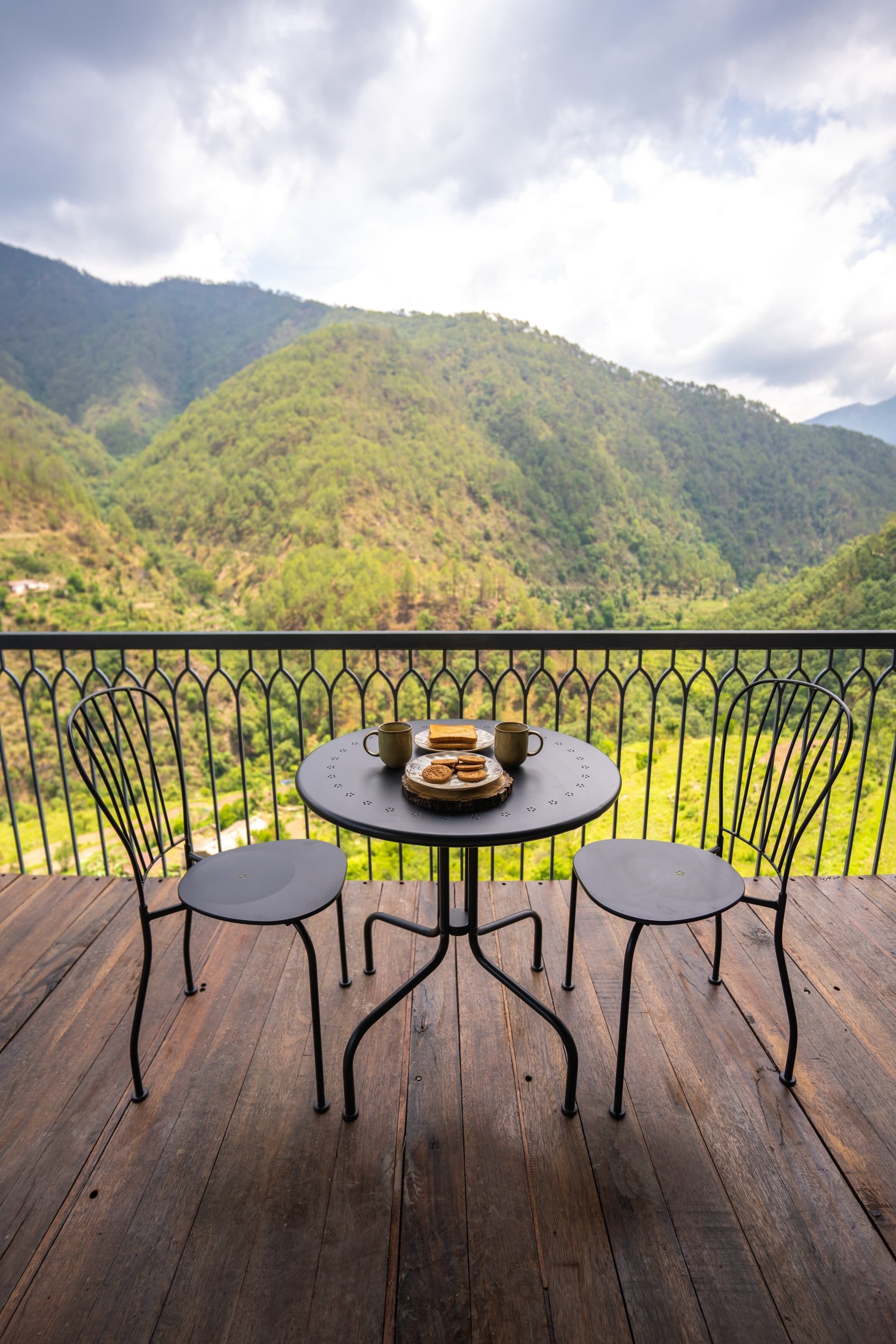 Um deck de madeira com uma mesa redonda e duas cadeiras, com vista para uma paisagem montanhosa.