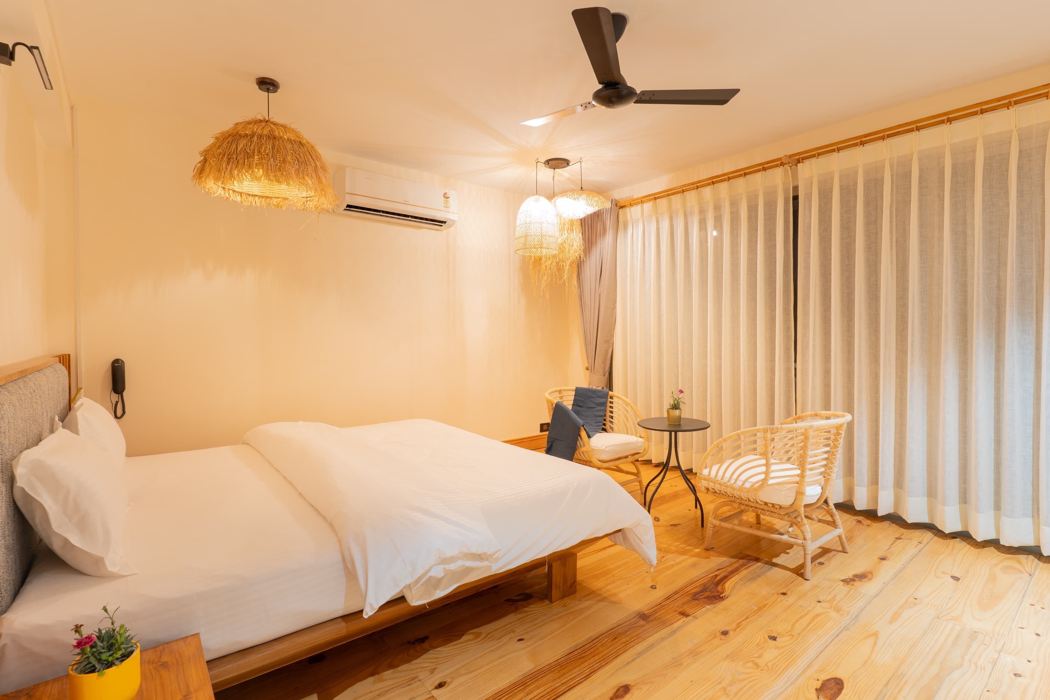 Quarto com cama, ventoinha no teto, luminária pendente, cadeira, mesinha e janela com cortinas.