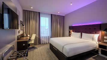 Quarto de hotel com cama, escrivaninha, cadeira, televisão e janela com cortinas.
