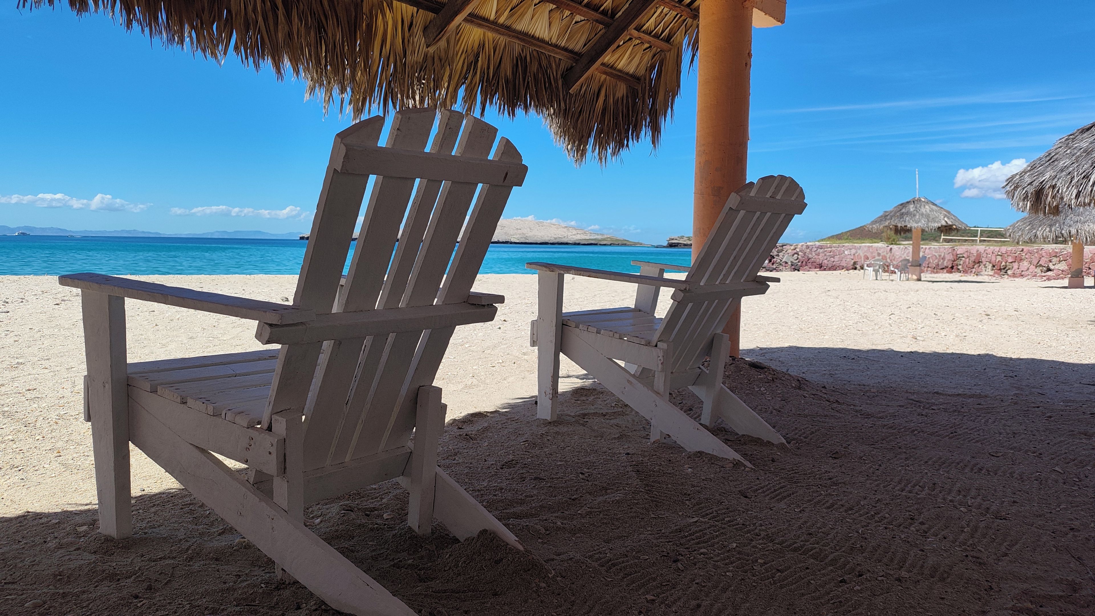 Duas cadeiras Adirondack sob um telhado de palha, colocadas numa praia arenosa com vista desimpedida para o oceano.