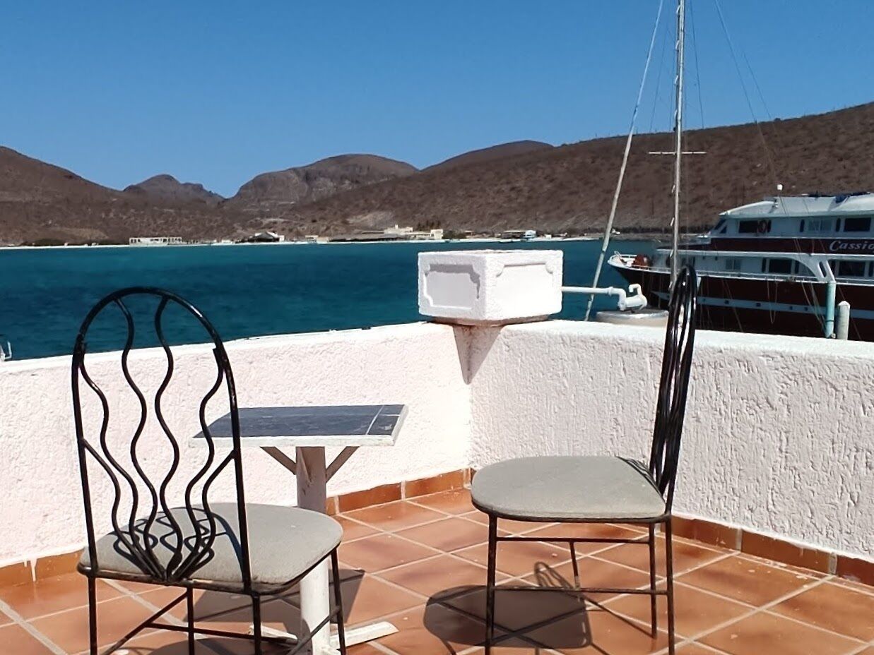 Terraço com duas cadeiras e uma mesa, com vista para um corpo de água e colinas.